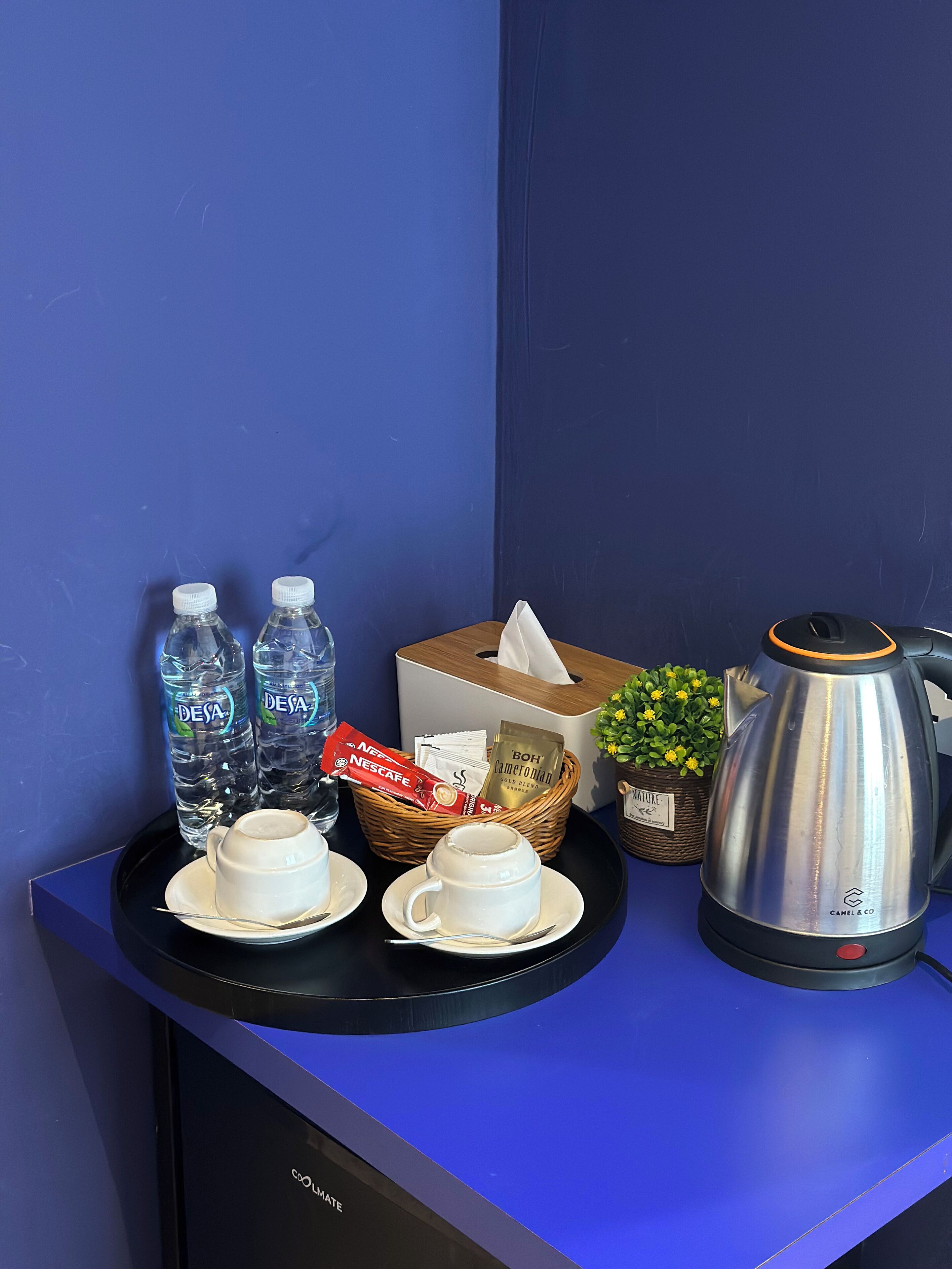 Uma bandeja com duas xícaras de café, duas garrafas de água, uma cesta com sachês de café, uma caixa de lenços de papel e uma chaleira em um balcão azul.