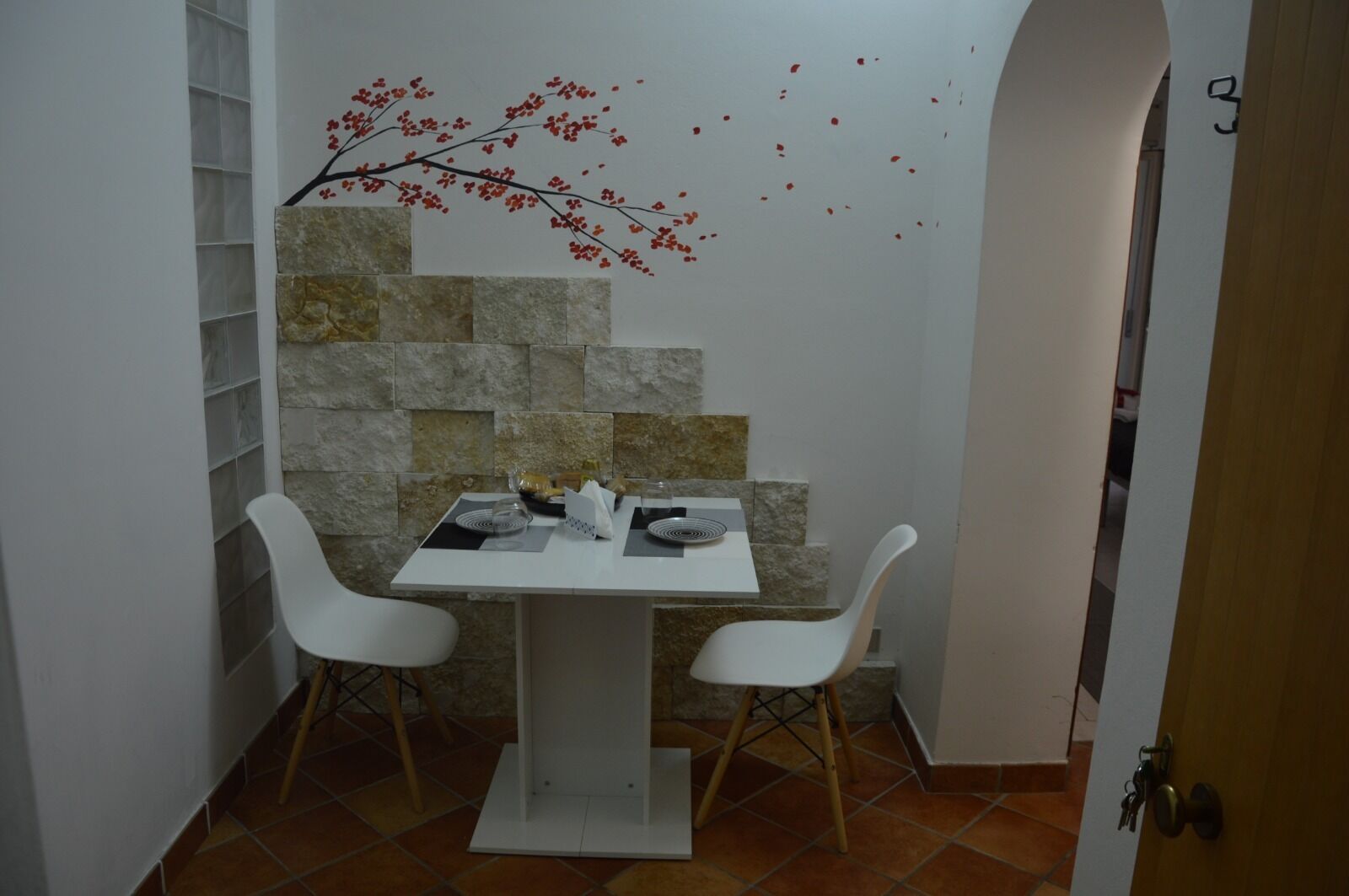 Uma sala de jantar com uma mesa branca e duas cadeiras brancas, uma parede de pedra com um detalhe de ramo, e um chão em mosaico.