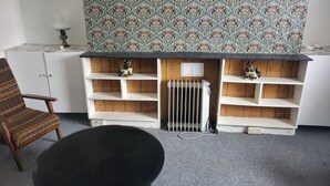 Um quarto com uma poltrona xadrez, uma mesa preta, um armário branco com prateleiras e um radiador.