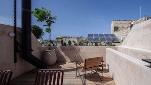 Um terraço no telhado com deck de madeira, cadeiras de metal, um vaso com planta e painéis solares ao fundo.