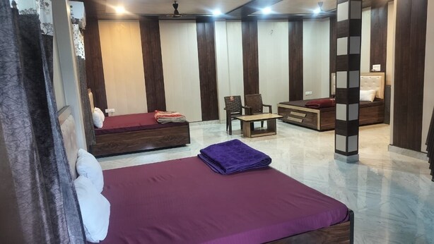 Quarto de hotel com uma cama grande, um sofá, uma mesinha e uma cadeira. Há cortinas nas janelas e um ventilador de teto.