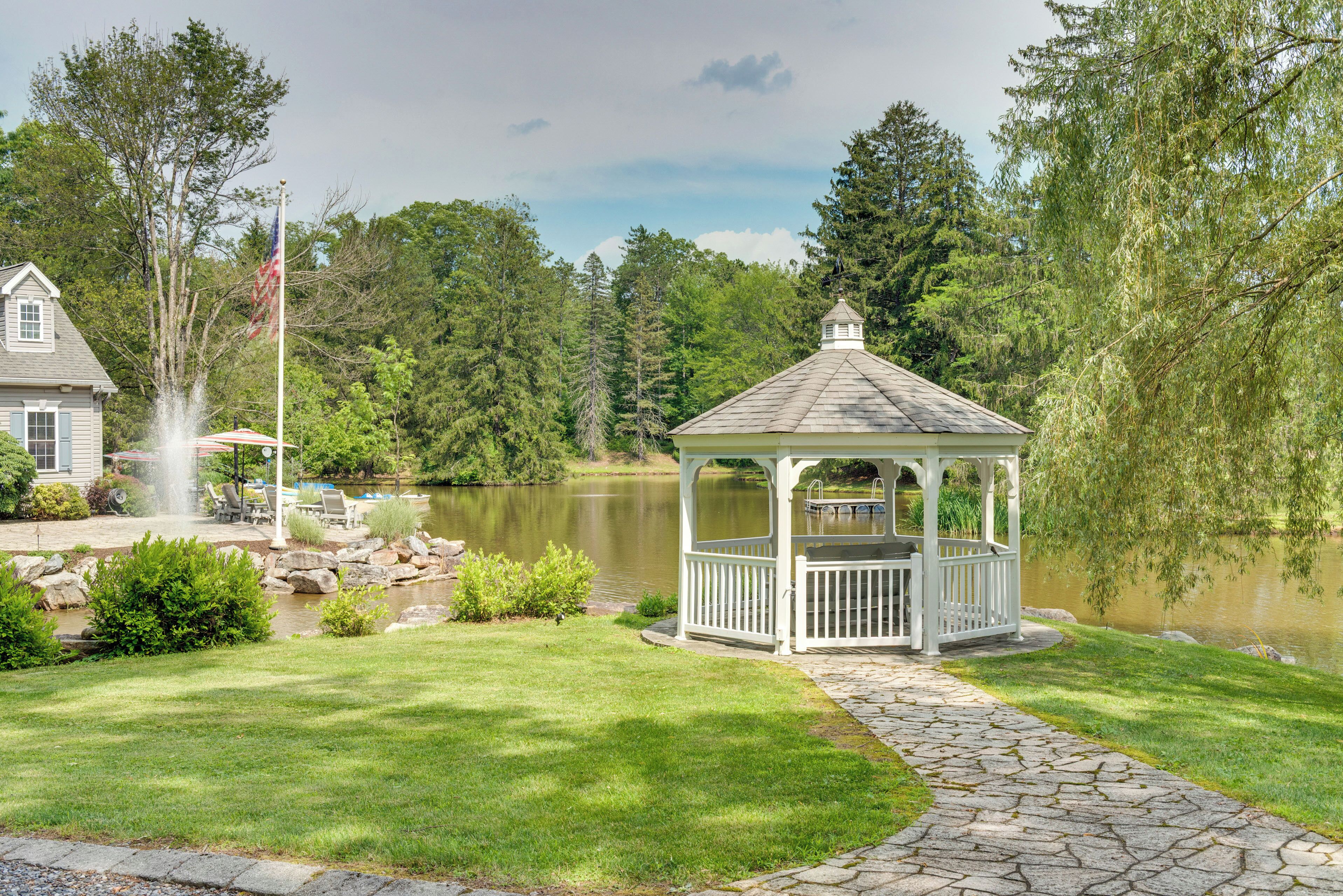Um gazebo com um caminho de pedra que leva a um lago, cercado por vegetação exuberante e uma casa com uma bandeira.