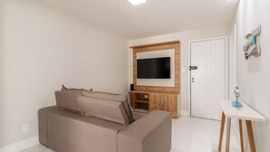 Uma sala de estar moderna com um sofá bege, uma TV de tela plana fixada em uma parede de madeira, uma pequena mesa com objetos decorativos e uma porta branca.