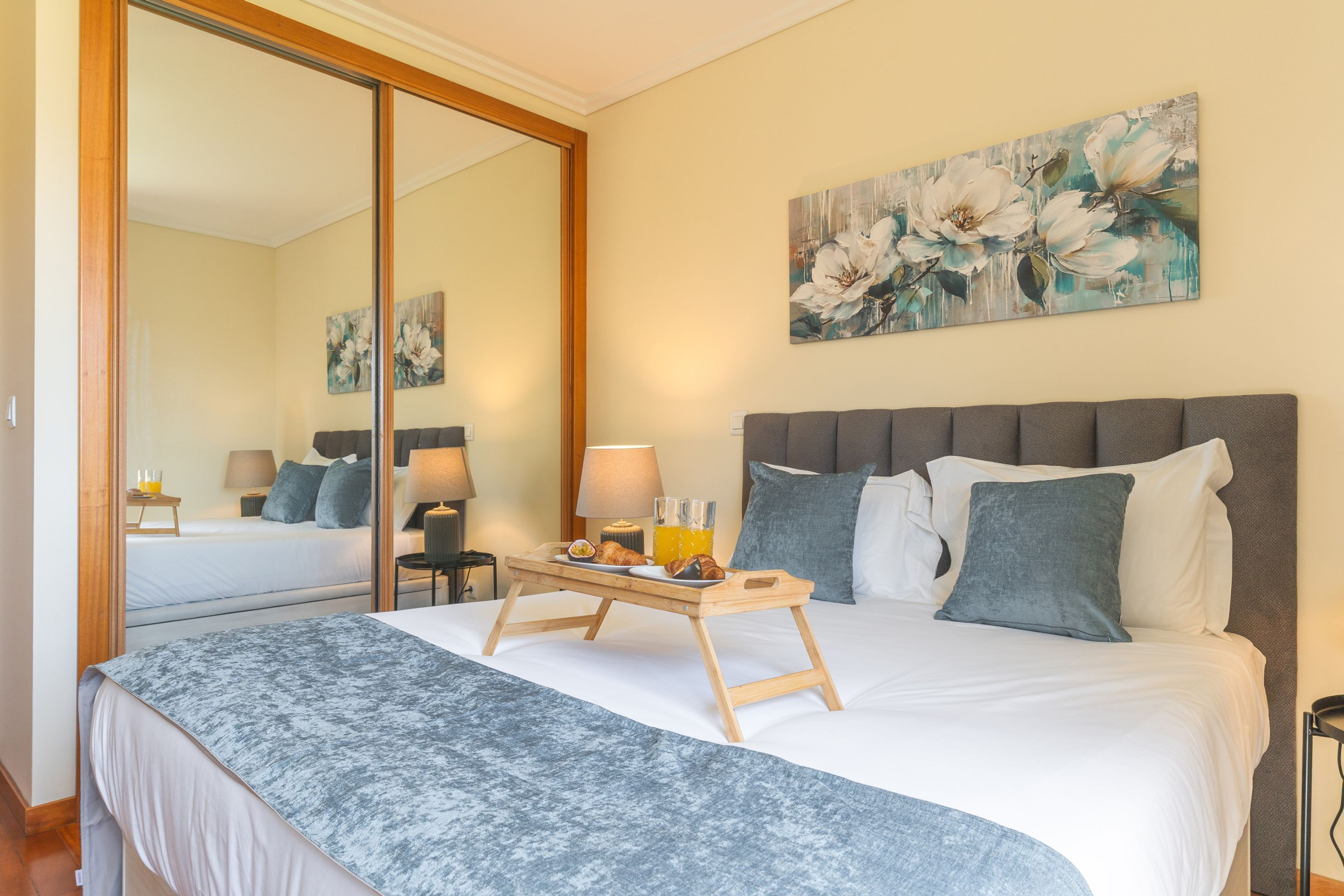 Quarto de hotel com uma cama grande, uma mesa de cabeceira com bandeja de café da manhã, um espelho e um quadro na parede.