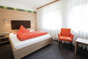 Quarto de hotel moderno com uma cama, uma cadeira laranja, cabeceira de madeira e uma janela grande com cortinas leves.