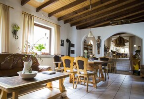 Uma sala de estar aconchegante com teto de madeira, uma área de refeições com cadeiras e mesa de madeira, e uma cozinha visível através de um arco.