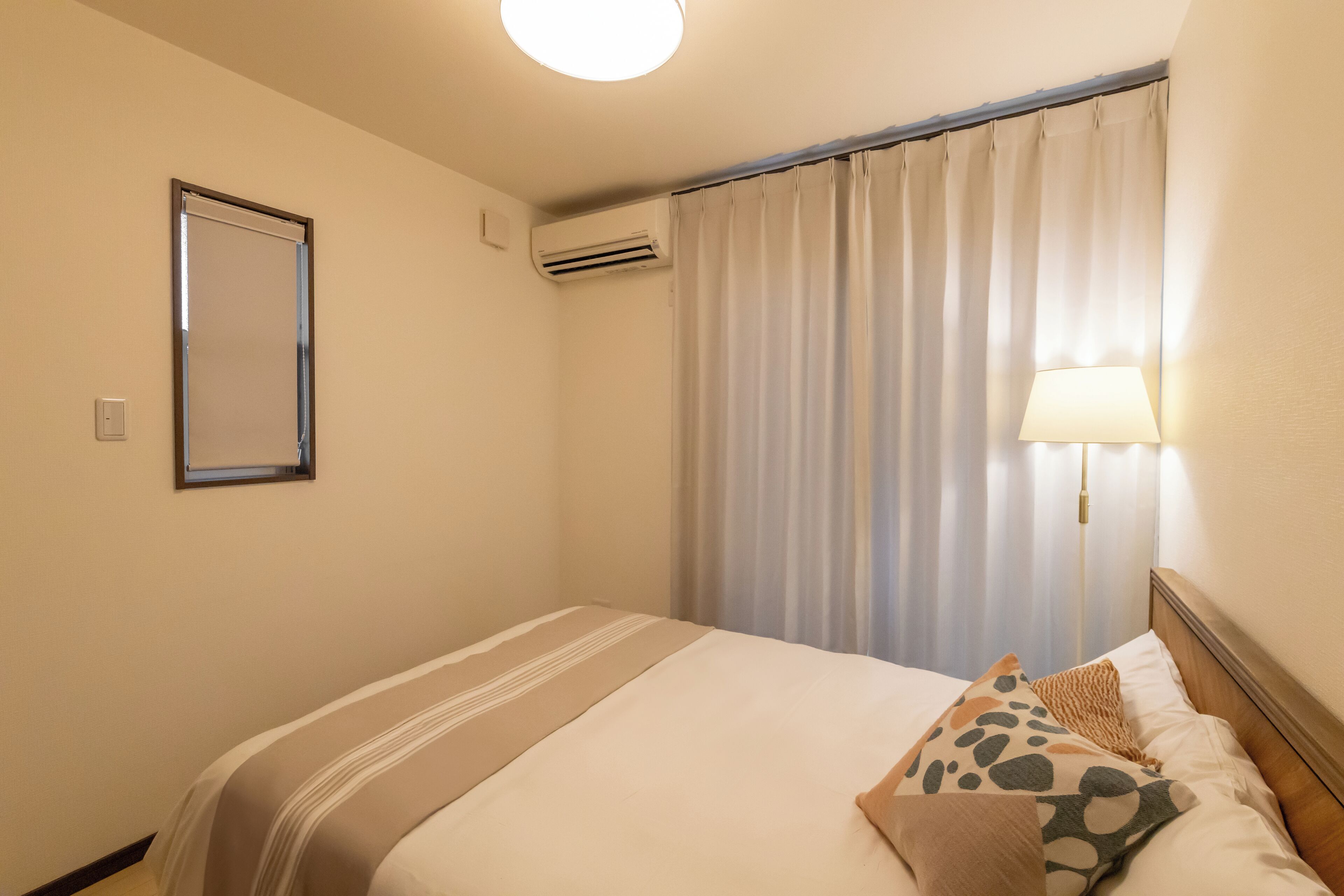 Quarto de hotel com cama, abajur, janela com cortinas, unidade de ar condicionado e espelho fixado na parede.