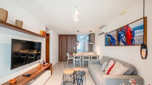 Uma sala de estar moderna com televisão de tela plana, um sofá cinza e uma área de jantar com cadeiras brancas.