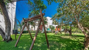 Um balanço de madeira numa área gramada, com árvores e um edifício ao fundo.