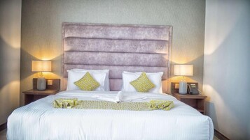 Cama bem arrumada com cabeceira, duas luminárias de cabeceira e mesa de apoio com telefone e vaso.
