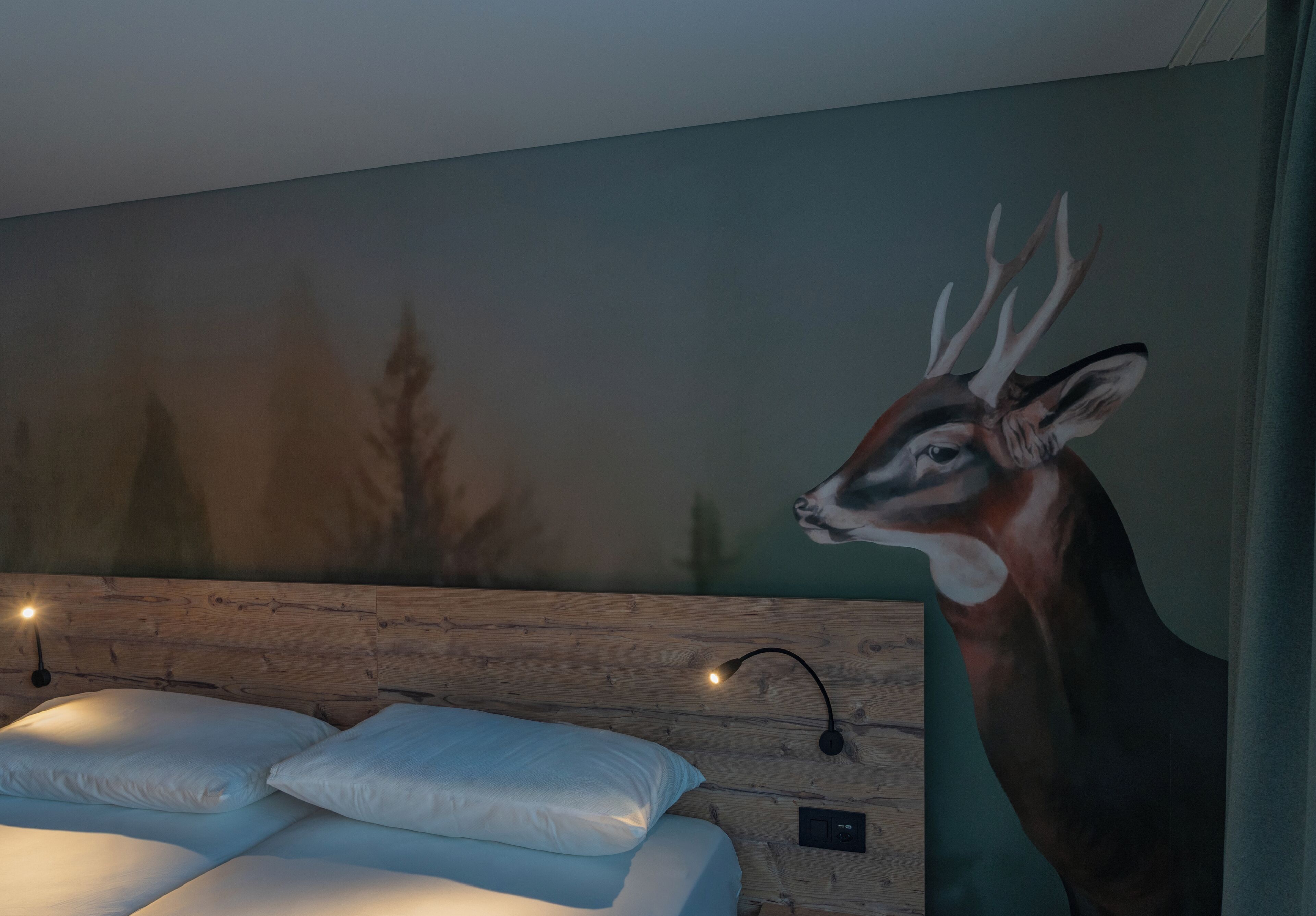 Quarto com cabeceira de madeira, duas almofadas brancas, um mural na parede com um veado e uma luz noturna.