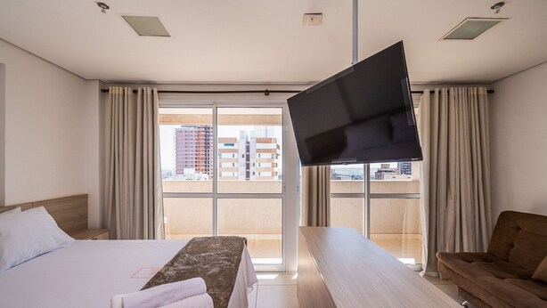 Quarto de hotel com cama, sofá, televisão fixada na parede e uma grande janela com cortinas.