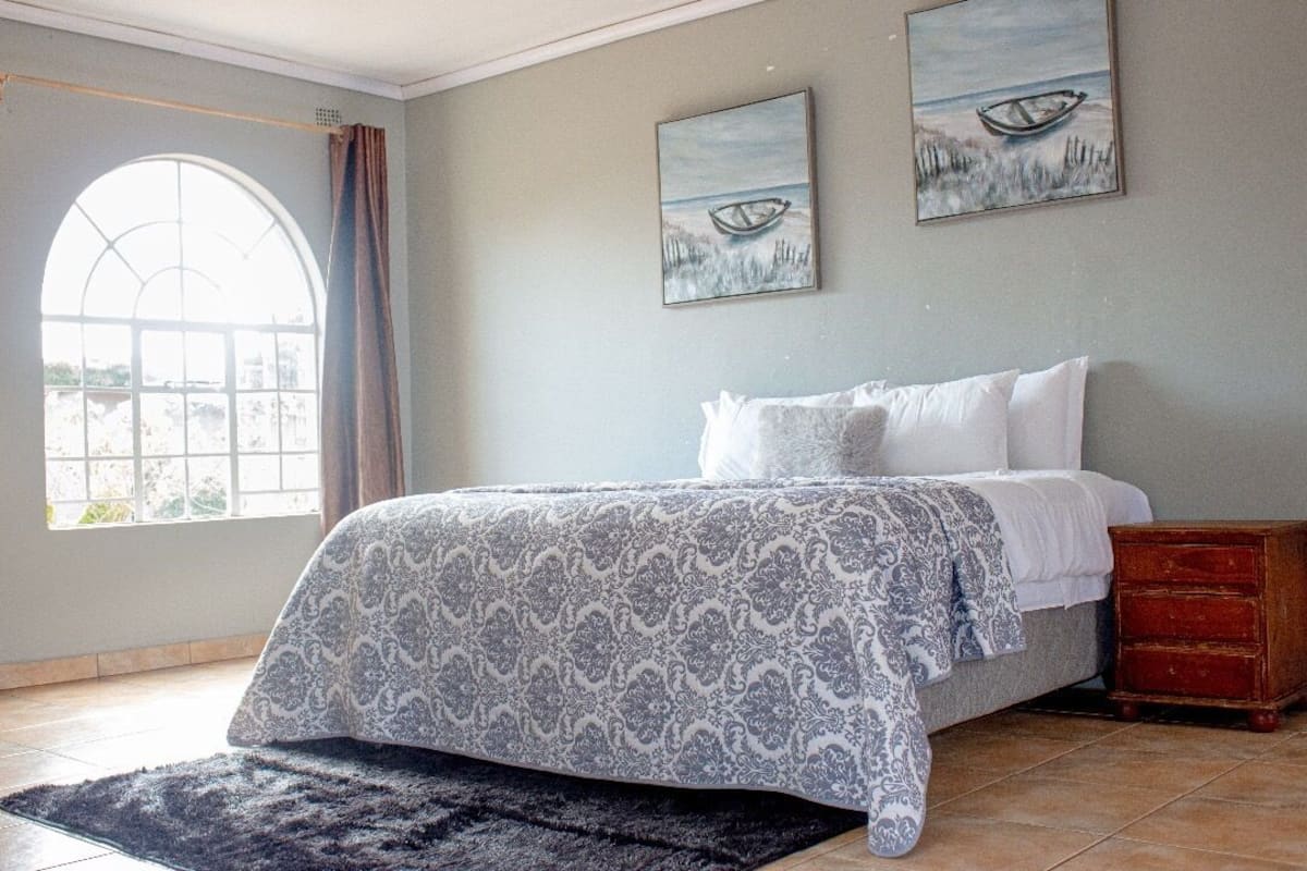 Quarto com uma grande janela, uma cama com roupa de cama estampada, uma mesinha de cabeceira de madeira e duas pinturas na parede.