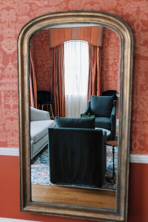 Quarto com espelho refletindo um sofá, poltrona, banquinho e uma janela com cortinas.