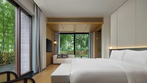 Quarto de hotel moderno com uma cama grande, um sofá, uma TV e vista para uma floresta através de grandes janelas.