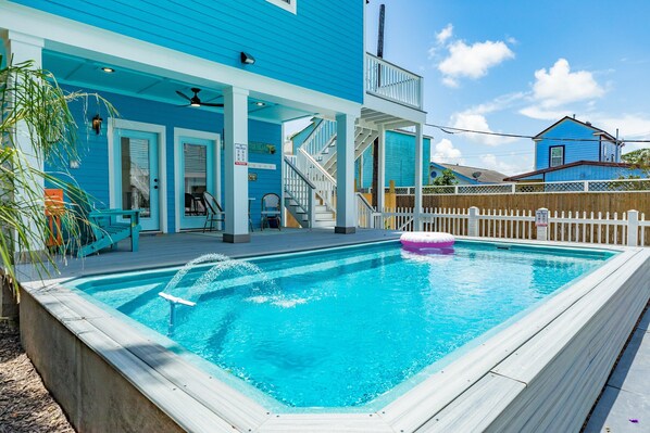Uma piscina com deck de madeira, área de estar coberta e uma casa azul ao fundo.