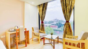 Quarto com cama, escrivaninha com chaleira, duas cadeiras, mesa pequena com planta e janelas amplas com cortinas.