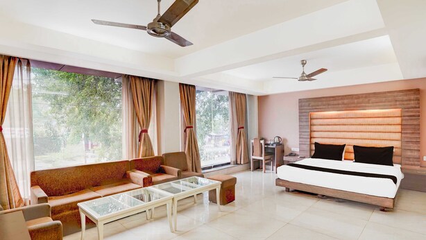 Quarto de hotel com sofá, mesa de centro de vidro, cama com cabeceira de madeira, ventilador de teto e grandes janelas com cortinas.