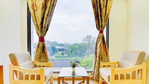 Quarto com duas cadeiras de madeira, uma mesa de vidro e vista para uma paisagem verde através de uma janela com cortinas estampadas.