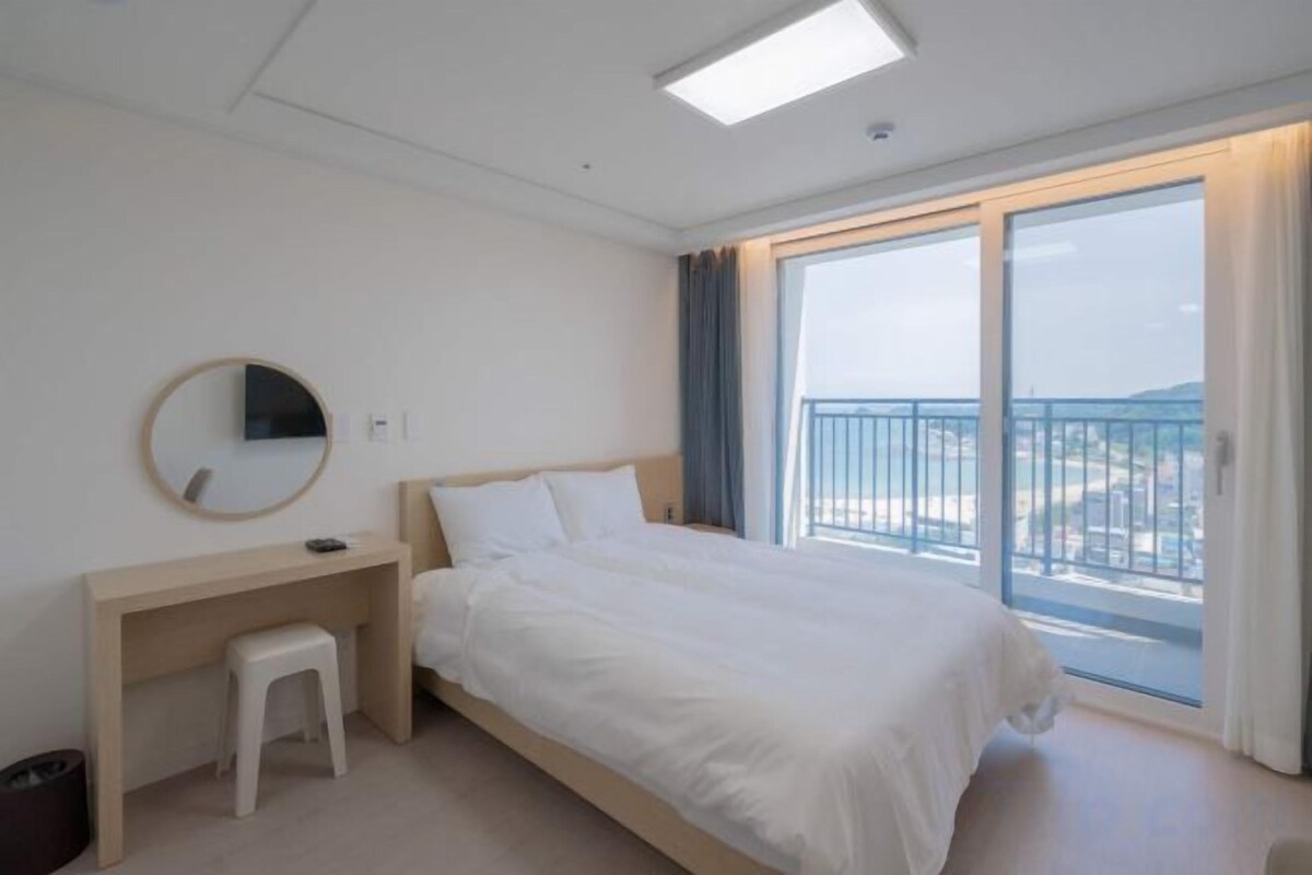 Quarto de hotel com cama, escrivaninha, cadeira, espelho e uma janela grande com vista.