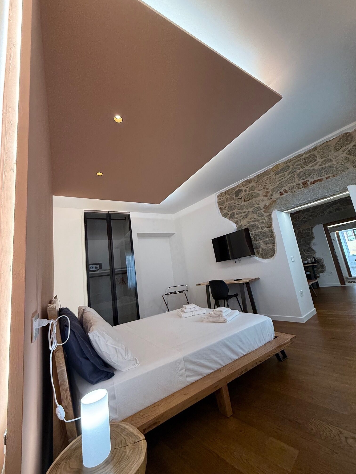 Quarto moderno com cama estilo loft, uma escrivaninha pequena, televisão fixada em uma parede de pedra e piso de madeira.