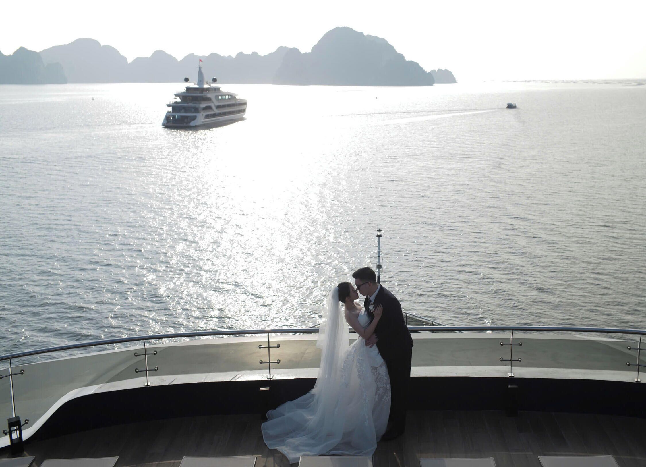 Um casal de noivos em um barco, com vista para uma grande extensão de água e formações terrestres ao longe.