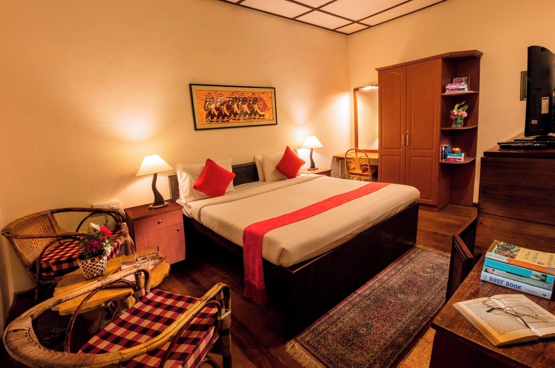 Quarto de hotel com cama, duas luminárias de cabeceira, guarda-roupa de madeira, pequena mesa com cadeira, estante e um quadro na parede.