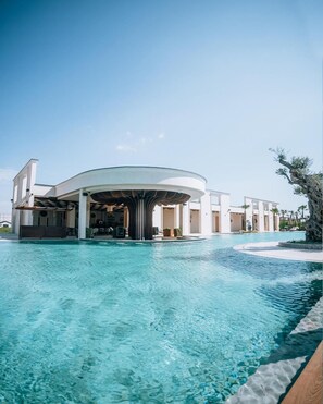 Um hotel moderno com uma piscina grande, área externa coberta e céu azul vibrante.