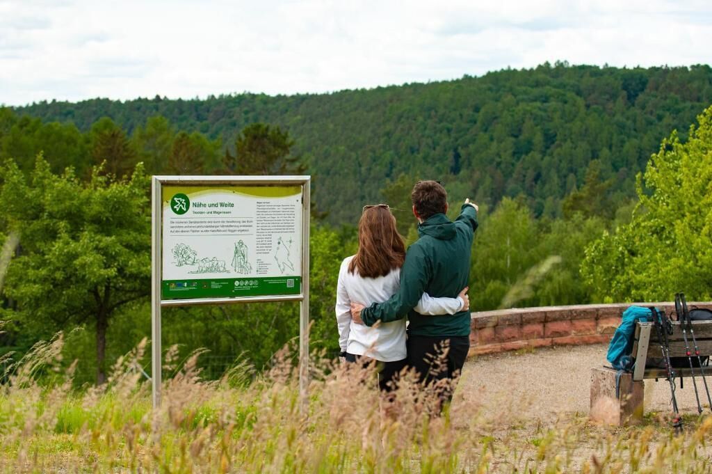 Um casal parado perto de um painel de sinalização, com uma paisagem natural ao fundo.