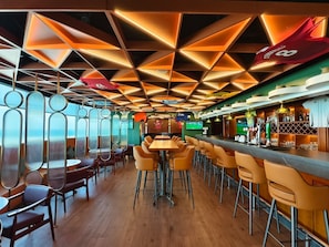Uma área de bar moderna com um balcão comprido, vários bancos e uma tela de televisão.