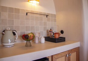 Uma cozinha com painel de azulejos, chaleira, tigela de frutas, xícaras empilhadas e cafeteira.