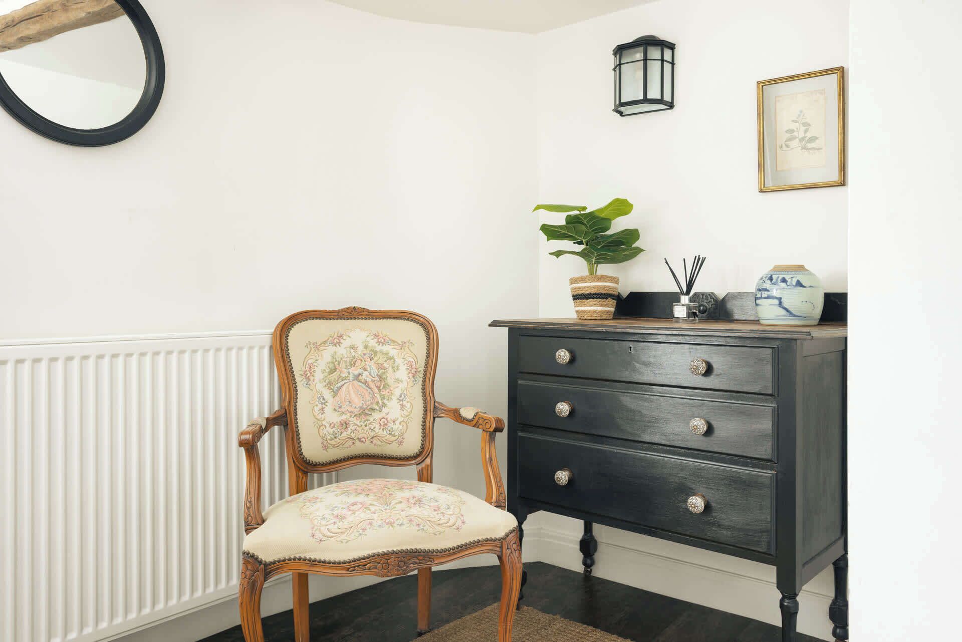 Quarto com cadeira de madeira com estofamento floral, cômoda de madeira escura com gavetas, planta em vaso, lanterna fixada na parede e ilustração botânica emoldurada.