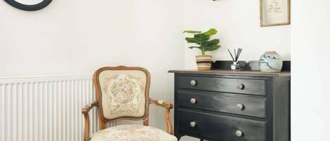 Quarto com cadeira de madeira com estofamento floral, cômoda de madeira escura com gavetas, planta em vaso, lanterna fixada na parede e ilustração botânica emoldurada.
