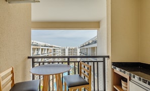 Uma varanda com uma mesa redonda e duas cadeiras, uma área de cozinha com bancada preta e vista para edifícios ao longe.