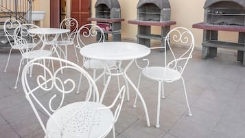 Um pátio com cadeiras e mesas de metal branco, duas fornalhas de pizza de pedra e uma escadaria que leva a uma porta.