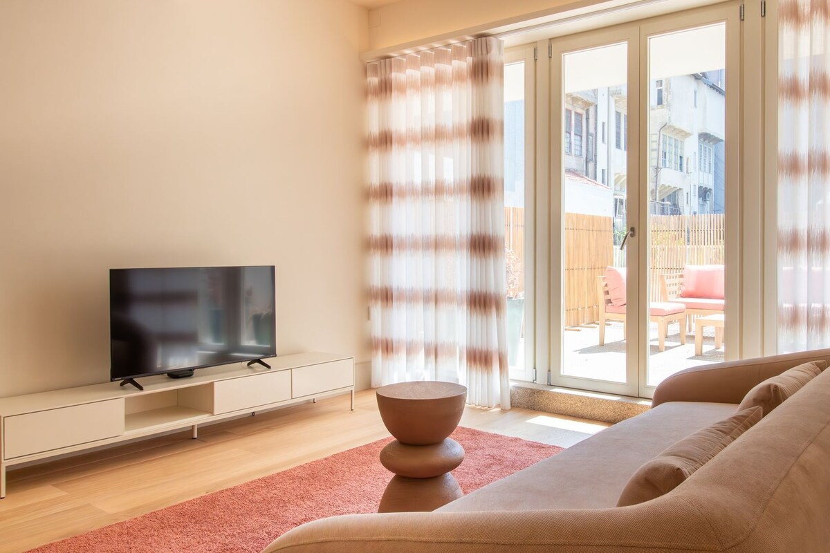 Uma sala de estar com televisão de tela plana em um suporte, um sofá bege, um tapete vermelho e uma grande porta de vidro deslizante que dá para uma área externa com deck de madeira e cadeiras.