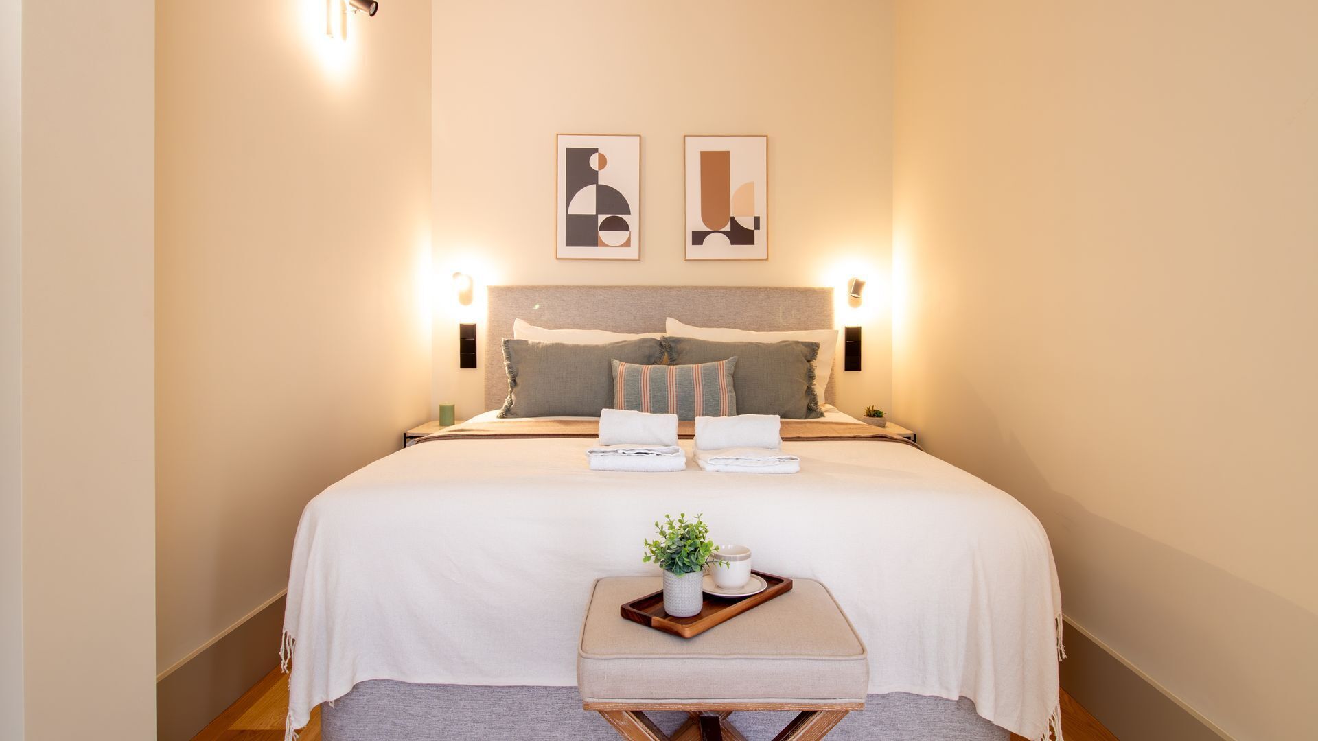 Cama bem arrumada com almofadas e um criado-mudo com luminária, além de uma bandeja com planta e xícara, e duas luzes fixadas na parede.