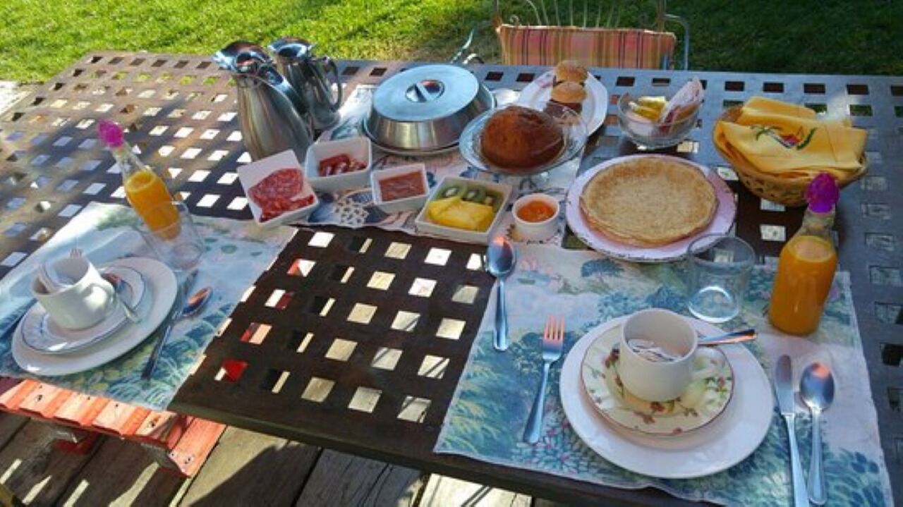 Mesa posta ao ar livre com diversos itens de café da manhã, incluindo um bule, xícaras, pratos e uma tigela de frutas.