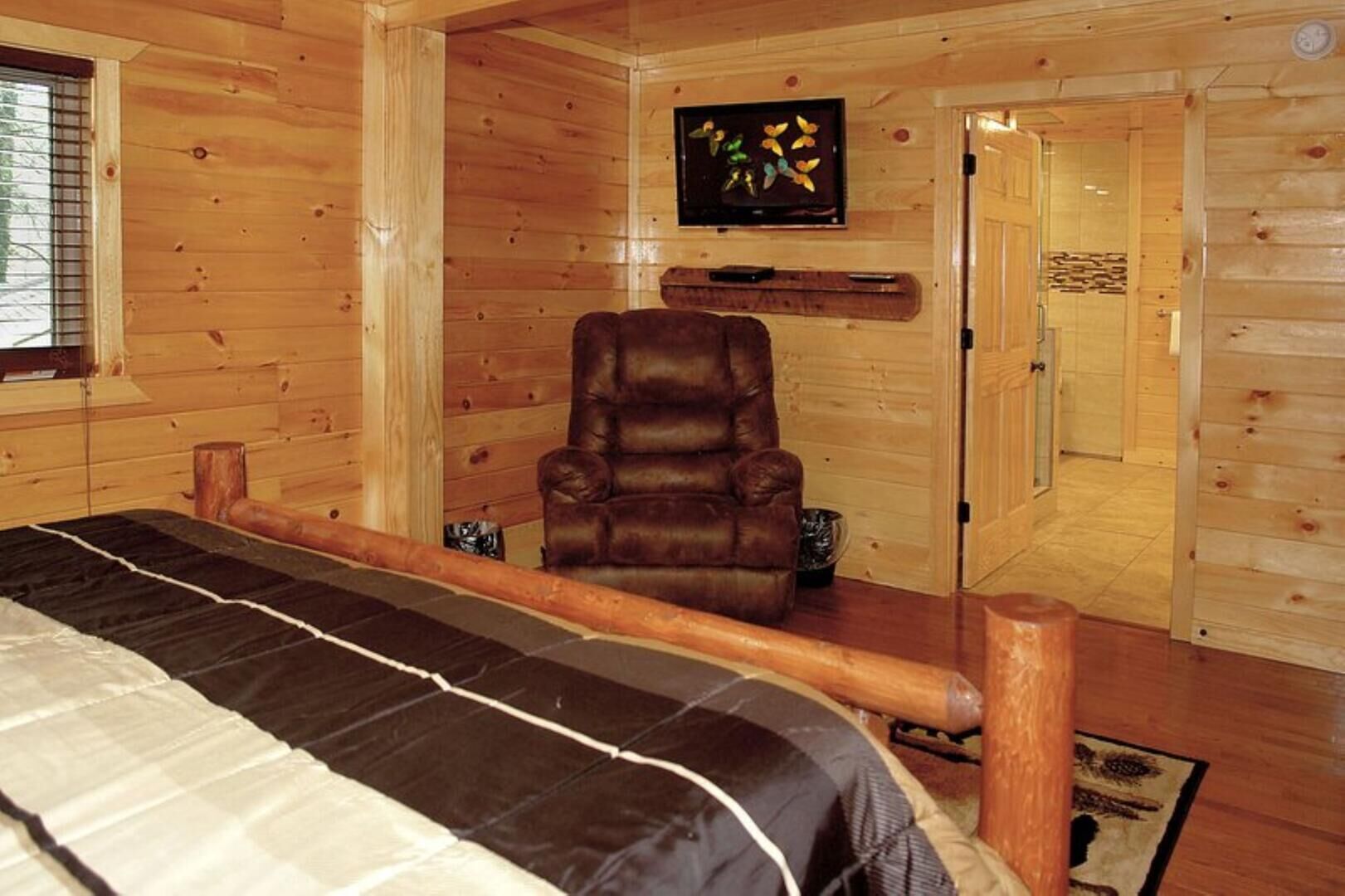 Um quarto de cabana de tronco com uma cama, uma poltrona de couro, uma lareira e uma obra de arte com borboletas emolduradas na parede.