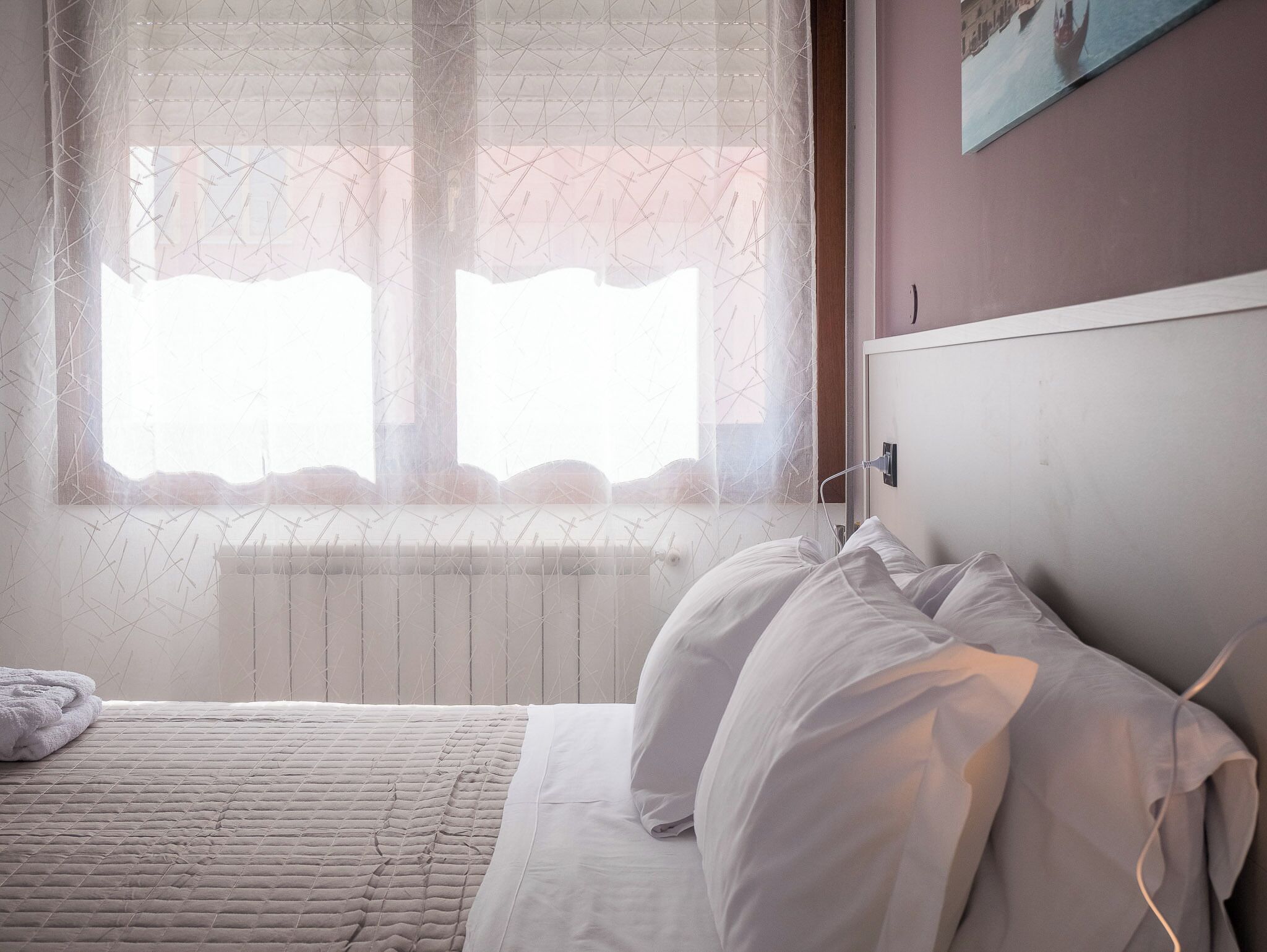 Quarto com cama, roupa de cama branca, janela com cortinas leves e um quadro na parede.