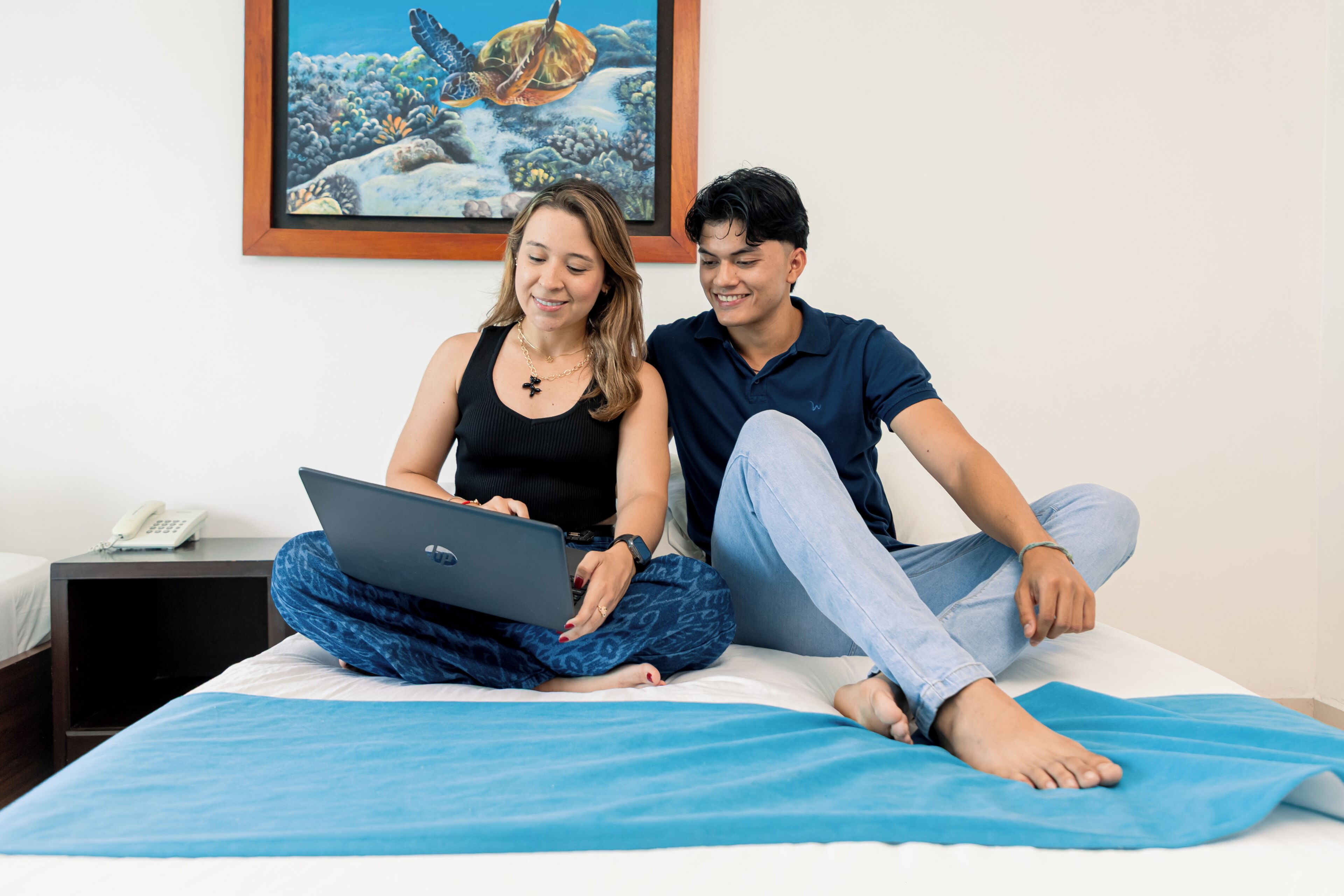 Duas pessoas sentadas na cama, com um laptop, um celular no criado-mudo e um quadro na parede.