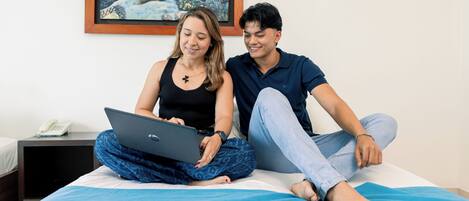Duas pessoas sentadas na cama, com um laptop, um celular no criado-mudo e um quadro na parede.