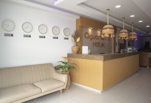 Uma área de recepção de hotel, com balcão de atendimento, um sofá e relógios nas paredes.
