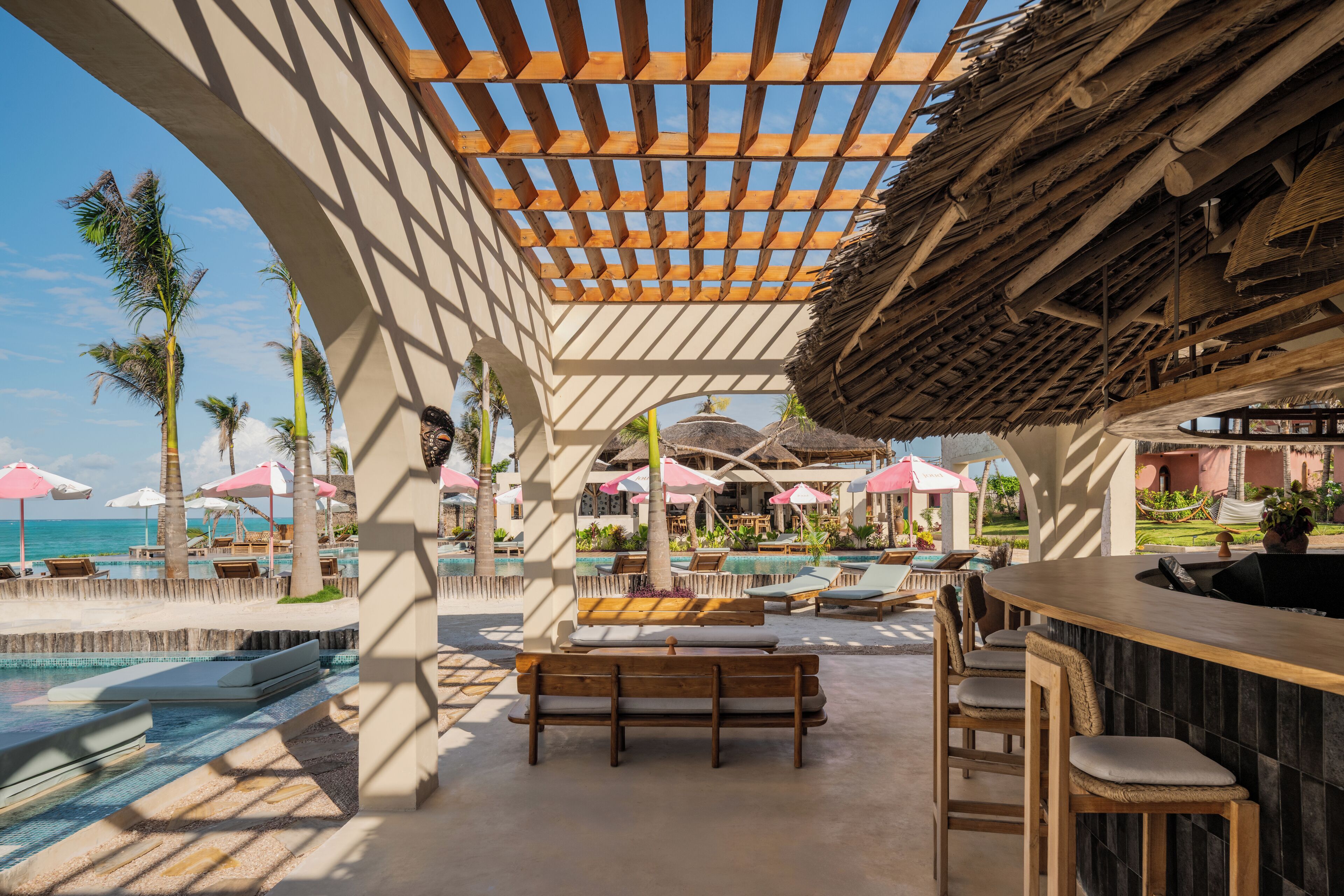 Uma área junto à piscina com um pergolado de madeira, espreguiçadeiras, um bar e vista para o oceano.