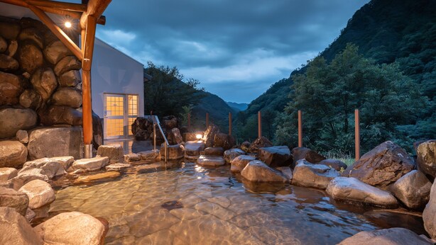 Uma onsen japonesa tradicional com banheira de pedra rodeada por pedras, estrutura de madeira e montanhas ao fundo.