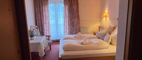 Quarto de hotel com uma cama grande, uma mesa pequena com uma cadeira e uma janela com cortinas.