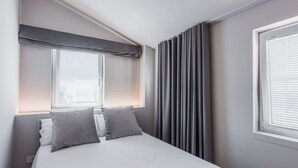 Quarto com cama, duas janelas com persianas, cortina e luminária sobre a cama.