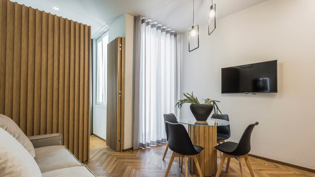 Uma sala de estar moderna com sofá, uma mesa de jantar com cadeiras, televisão na parede e um painel de madeira.