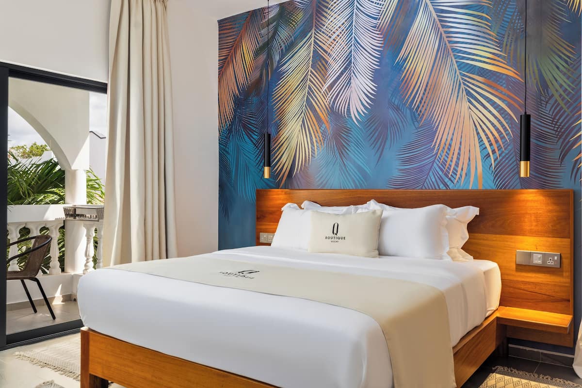Quarto de hotel com cama, cabeceira de madeira, parede com padrão de folha de palmeira, varanda com cadeira e luminária suspensa.