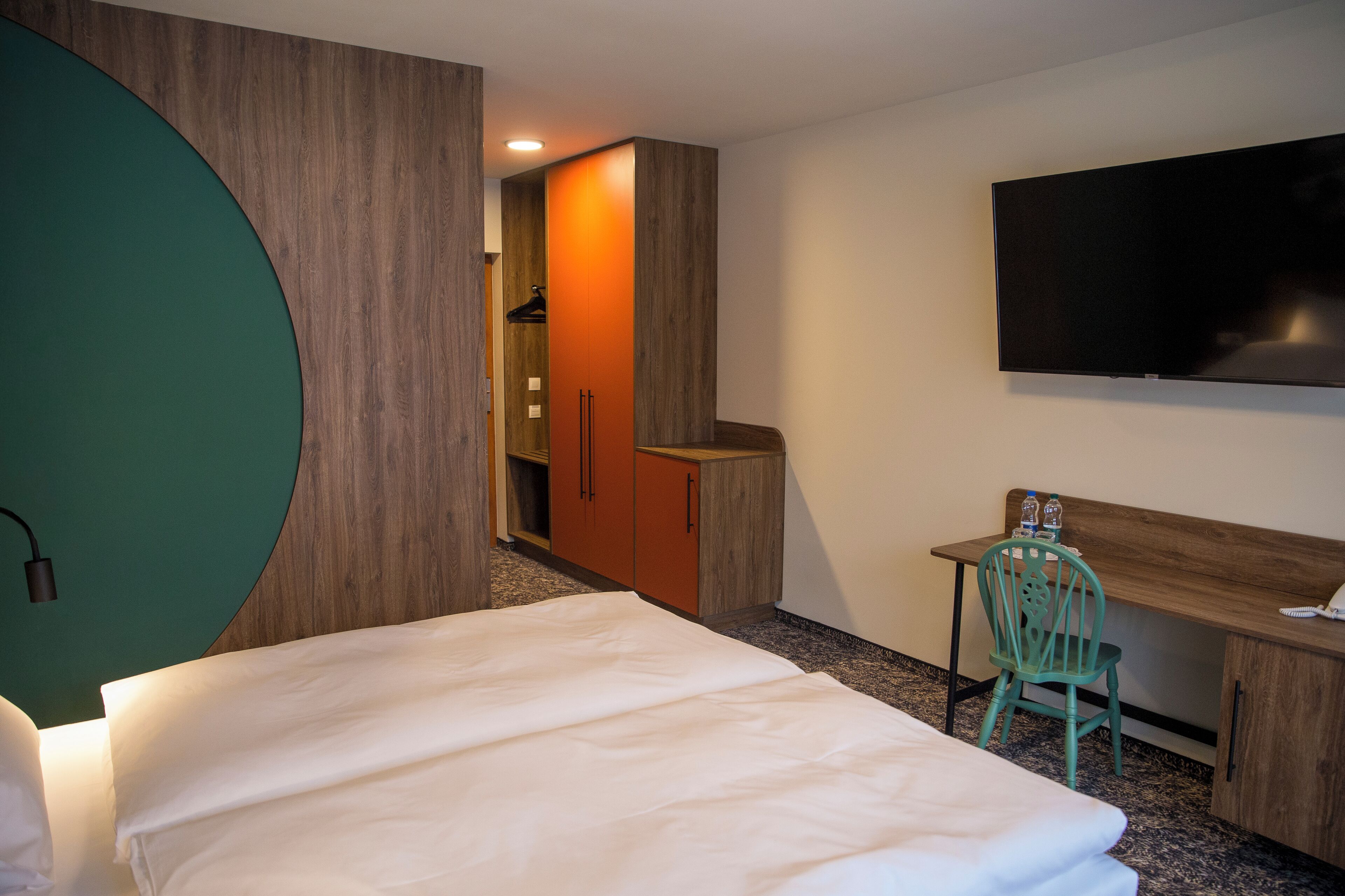 Quarto de hotel com uma cama, uma secretária, uma televisão e um guarda-roupa.